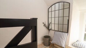 Quarto com uma cadeira preta, um banquinho de madeira, uma almofada estampada, um balde de metal e um espelho grande na parede.