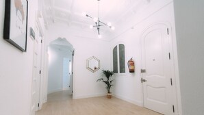 Um corredor com paredes brancas, piso de madeira, planta em vaso, quadro e extintor de incêndio.