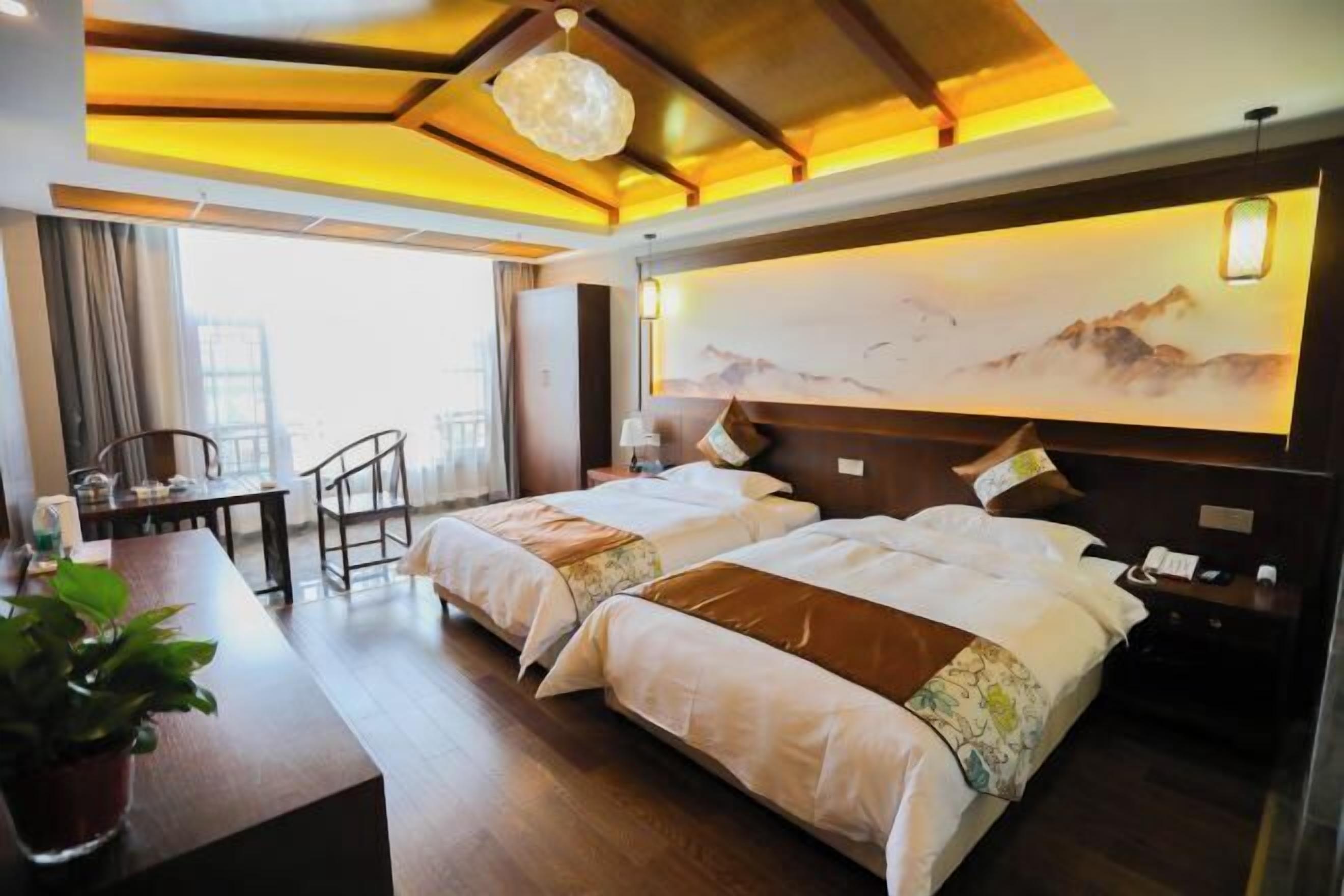 Quarto de hotel com duas camas, uma escrivaninha com cadeira, uma janela grande e um quadro na parede.