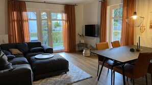 Uma sala de estar moderna com um sofá, uma mesa de jantar com cadeiras, uma televisão de tela plana na parede e grandes janelas com cortinas.