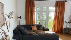 Uma sala de estar moderna com sofá seccional, mesa de centro redonda, mesa lateral e luminária de chão. Há uma janela grande com cortinas, um radiador e vista para o verde lá fora.