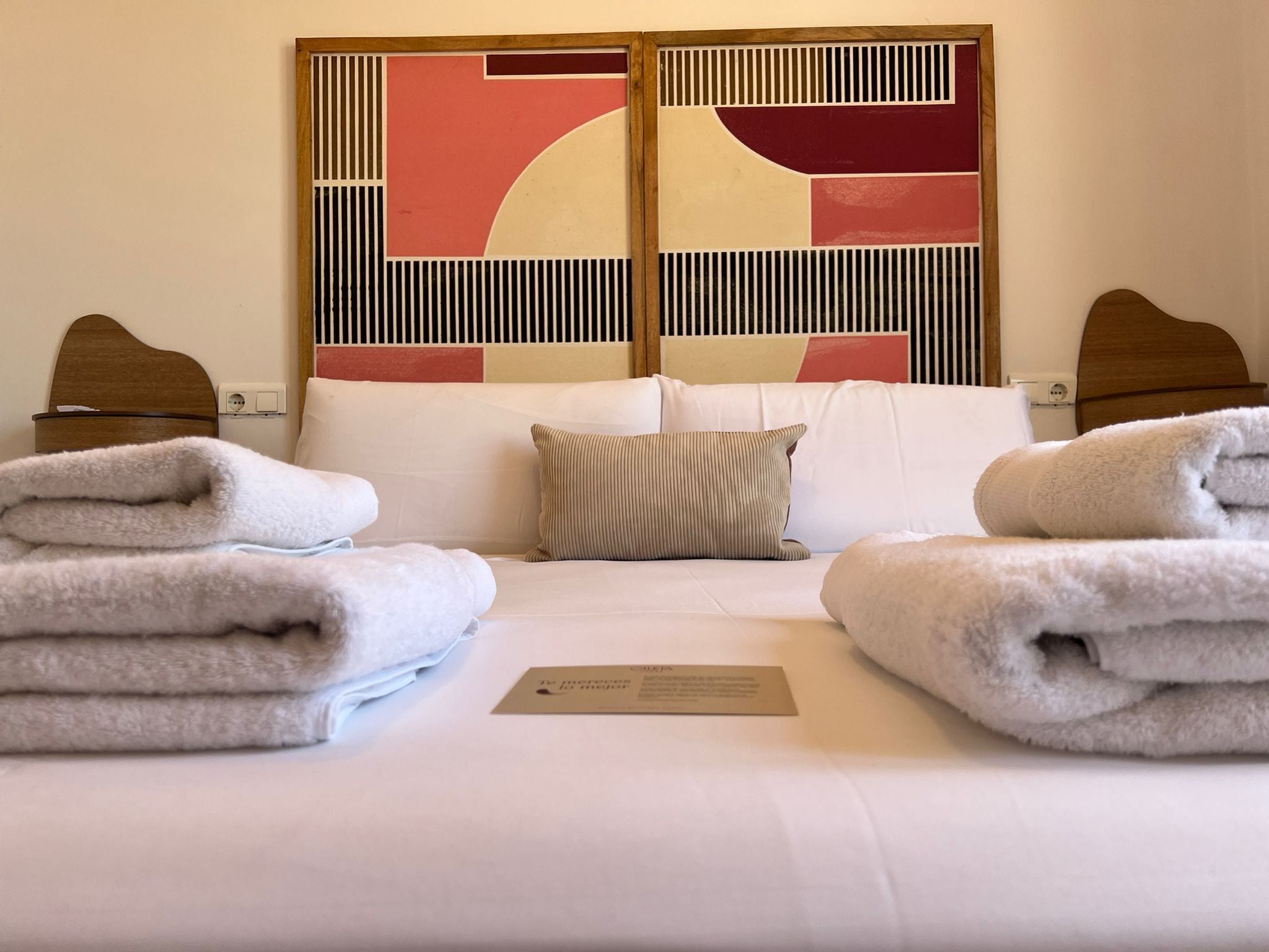 Quarto de hotel com cama, almofadas, toalha dobrada e uma placa sobre a cama.