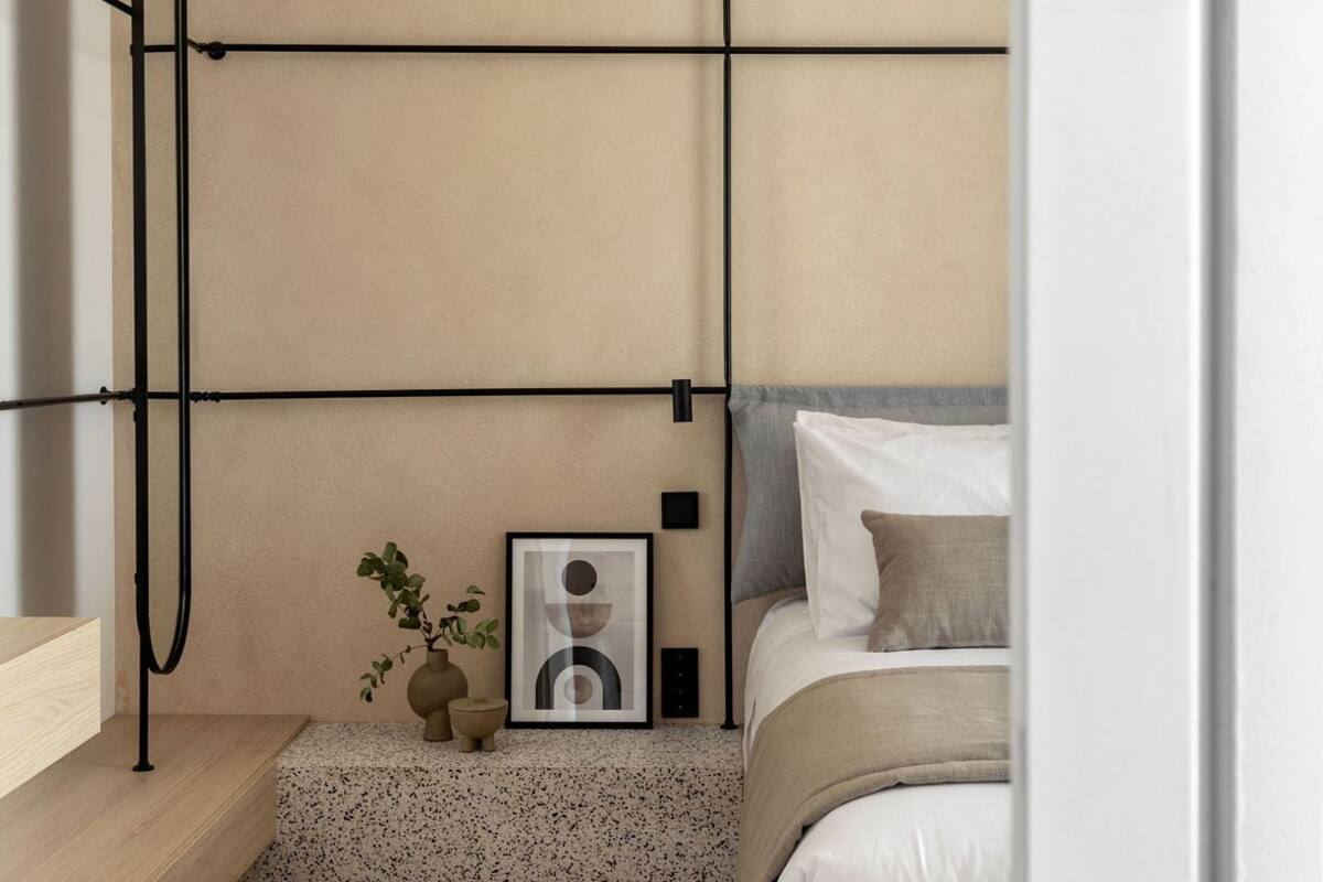 Quarto moderno com piso de madeira em padrão herringbone, cama com estrutura de madeira, cabeceira com grade de metal preto e uma pequena mesa de cabeceira com um vaso e um quadro emoldurado.