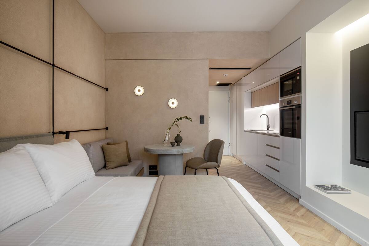 Quarto de hotel moderno com uma cama grande, uma mesa redonda pequena, uma cadeira e um espaço com cozinha compacta.