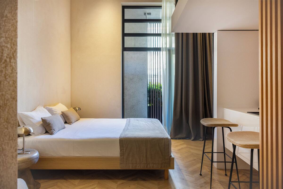 Quarto moderno com cama, mesas de cabeceira, janela com cortinas e vista para o verde lá fora.