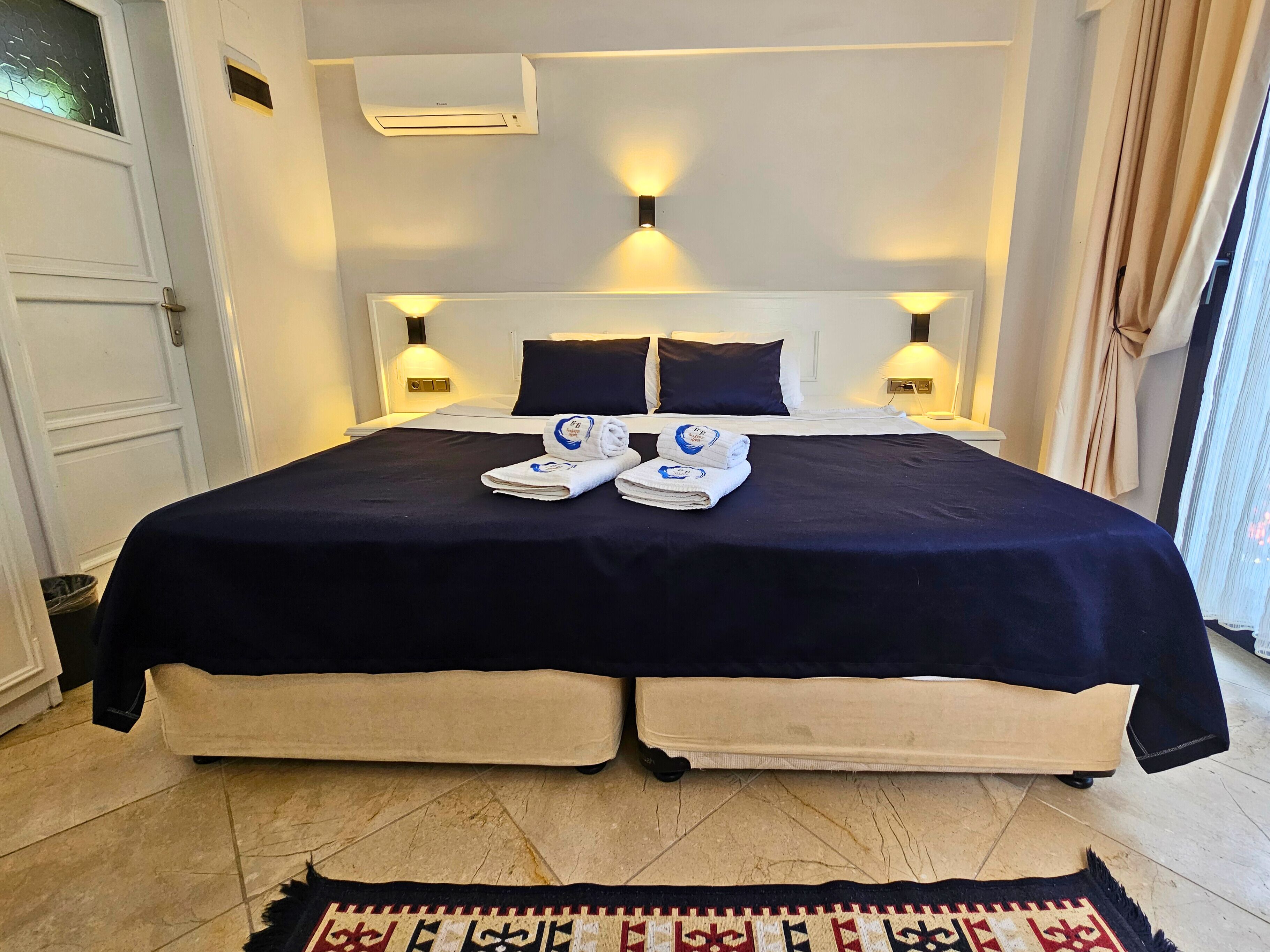 Quarto de hotel com uma cama grande, duas mesas de cabeceira, ar condicionado fixado na parede e uma janela com cortinas.