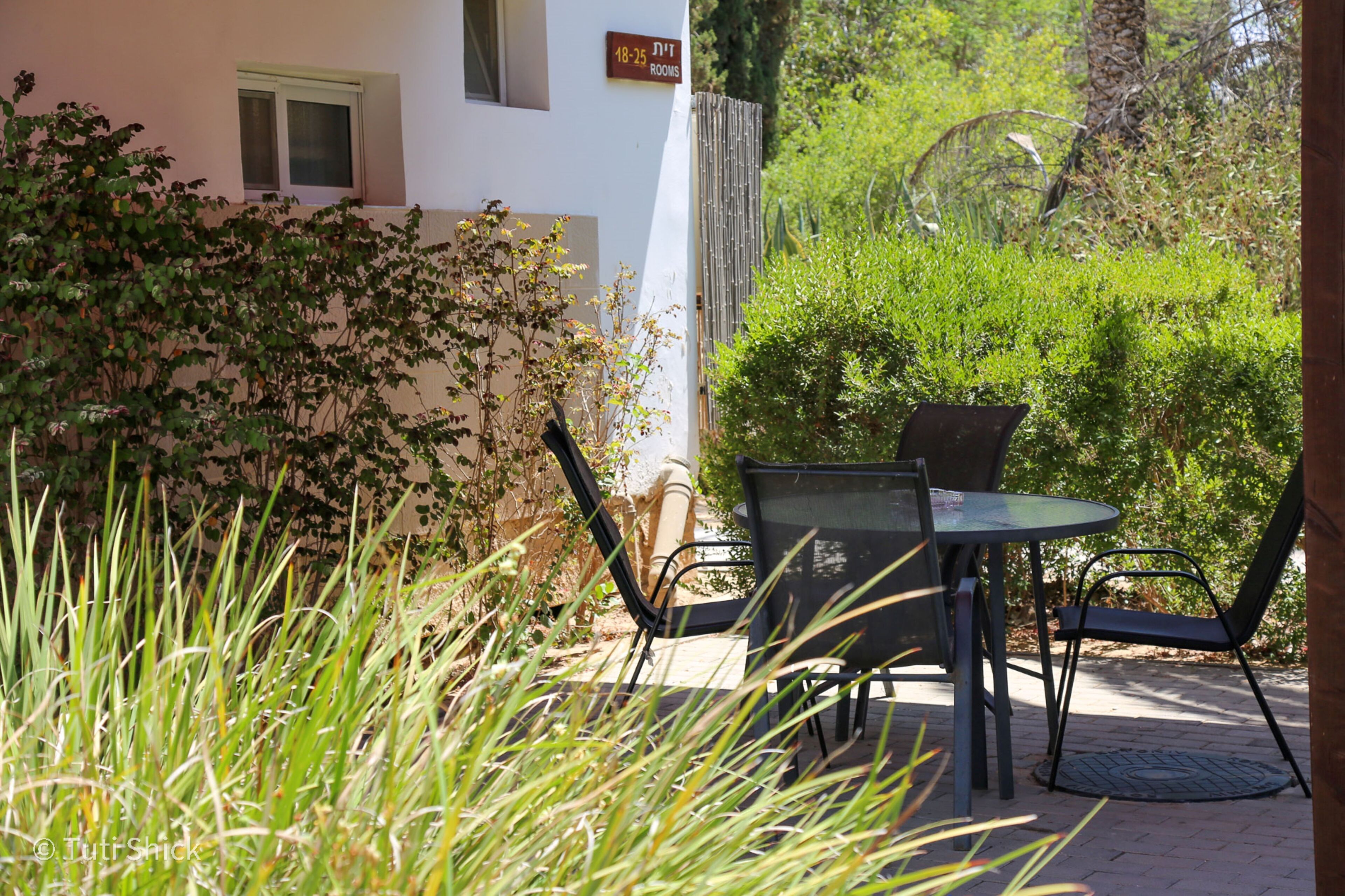 Um pátio com mesa e cadeiras, cercado por vegetação e um edifício com placa indicando quartos disponíveis.