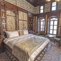 Quarto tradicional com parede de pedra, uma cama grande com roupa de cama estampada e uma pequena mesa com cadeiras.