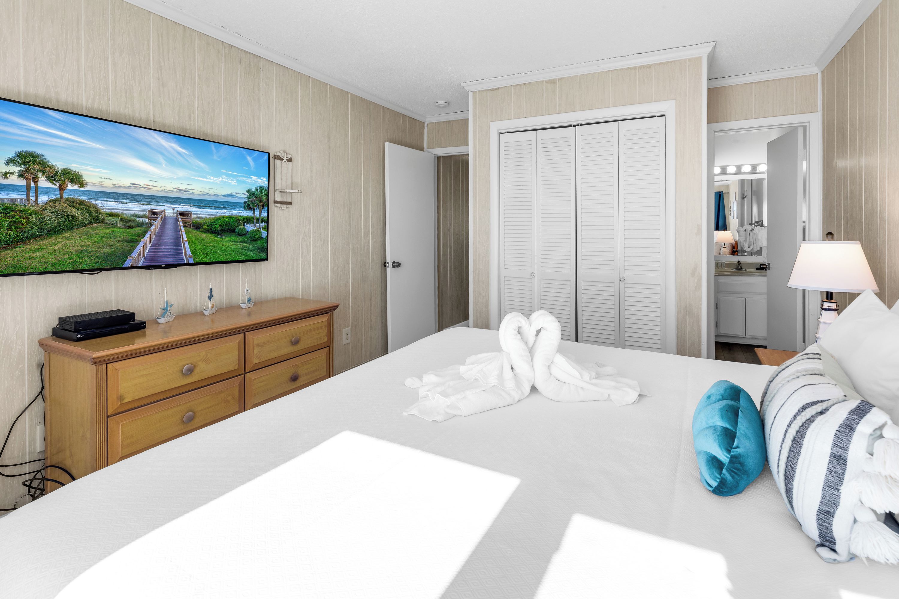 Quarto de hotel com cama, televisão, cómoda de madeira, armário e espelho de casa de banho.