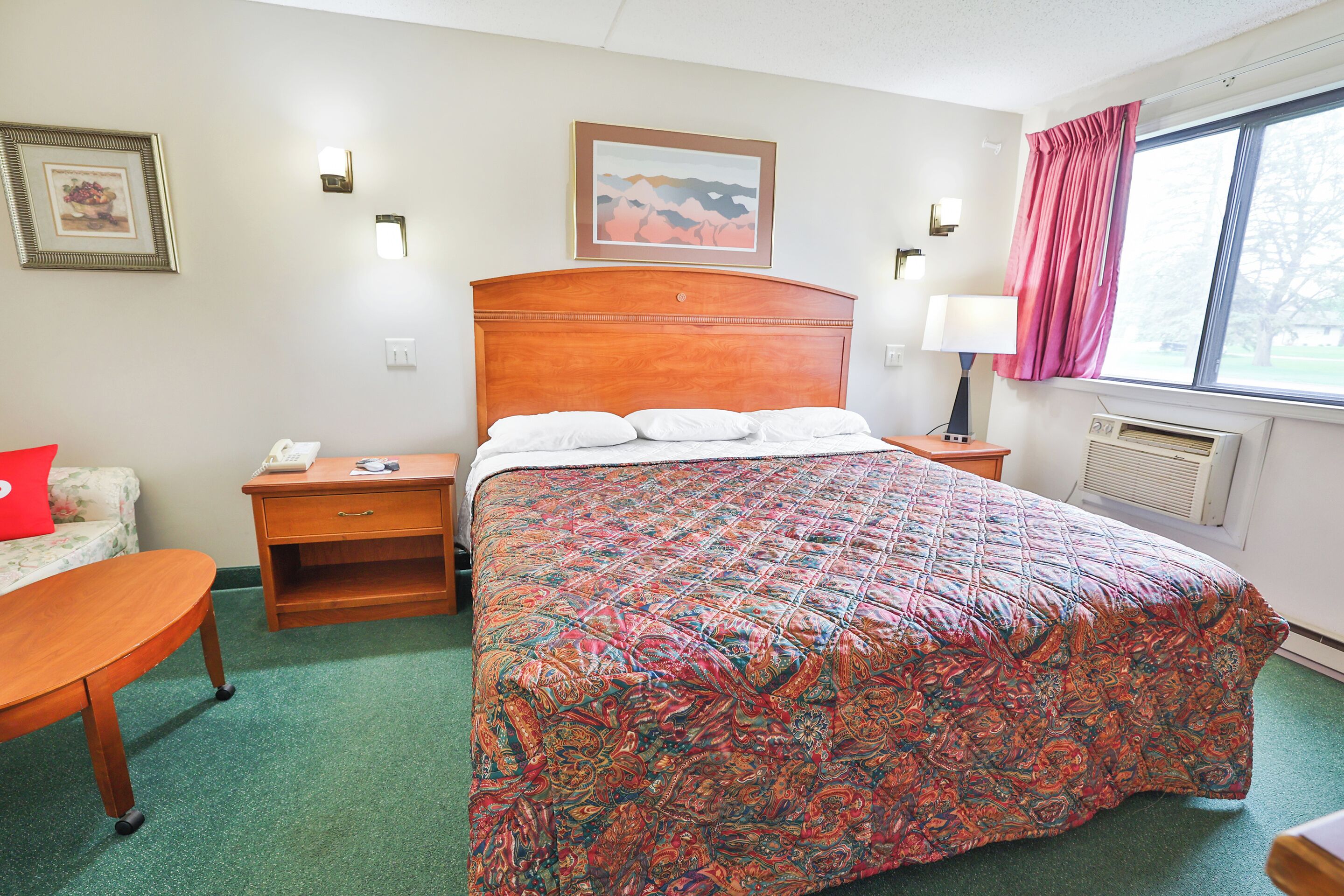 Quarto de hotel com cama, mesas de cabeceira, escrivaninha e cadeira. Há uma janela com cortinas, uma luminária na parede e um quadro emoldurado acima da cama.