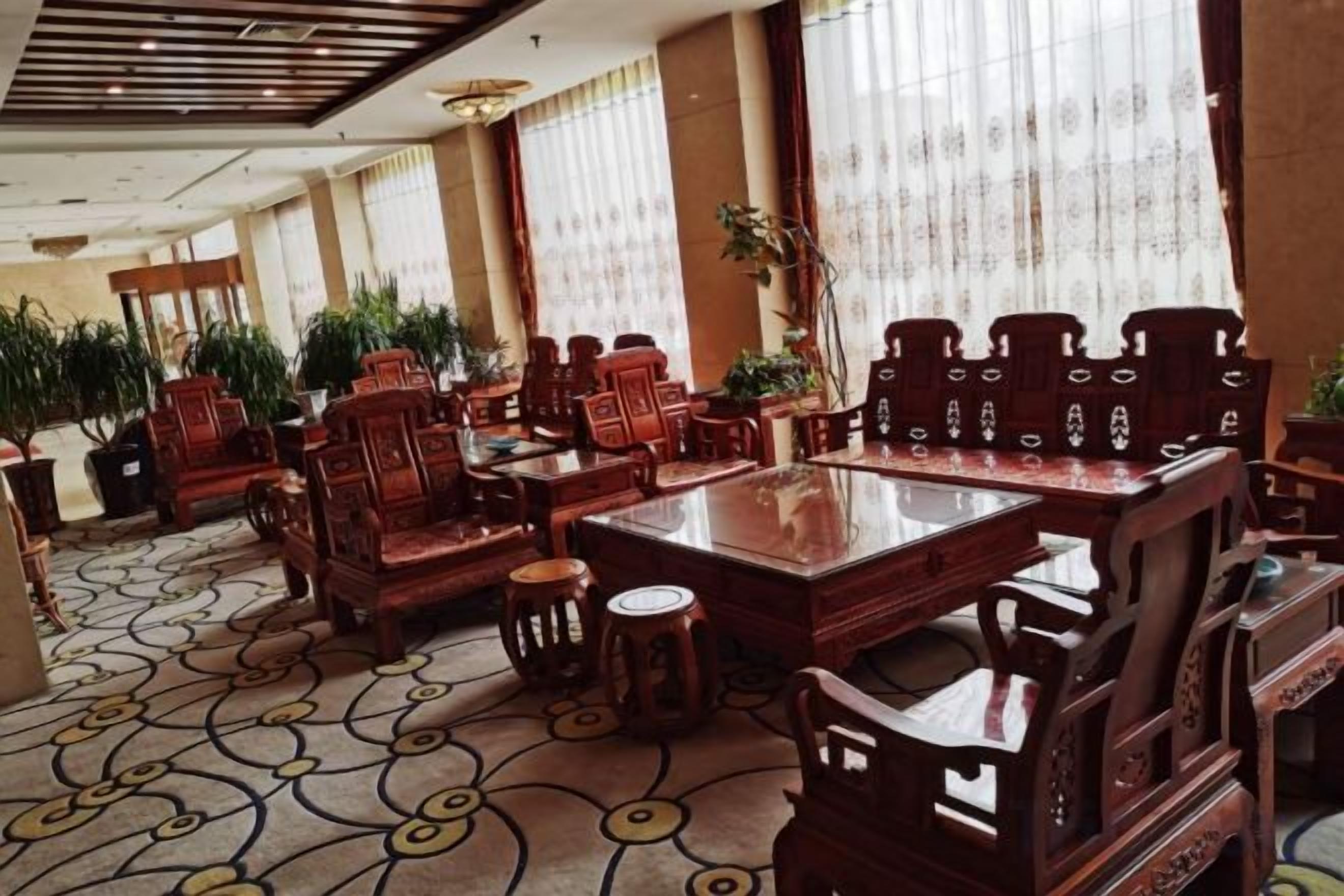 Área de refeições de hotel com mobiliário de madeira, carpete estampado e grandes janelas com cortinas.