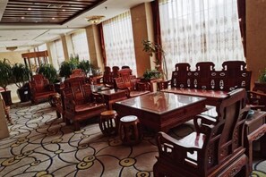 Área de refeições de hotel com mobiliário de madeira, carpete estampado e grandes janelas com cortinas.