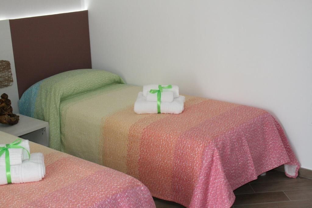 Quarto de hotel com duas camas, cada uma com um edredom de cor diferente, e um par de toalhas dobradas com um laço verde.