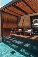 Área de piscina com teto de ripas de madeira, duas espreguiçadeiras e porta de vidro que dá para o exterior.