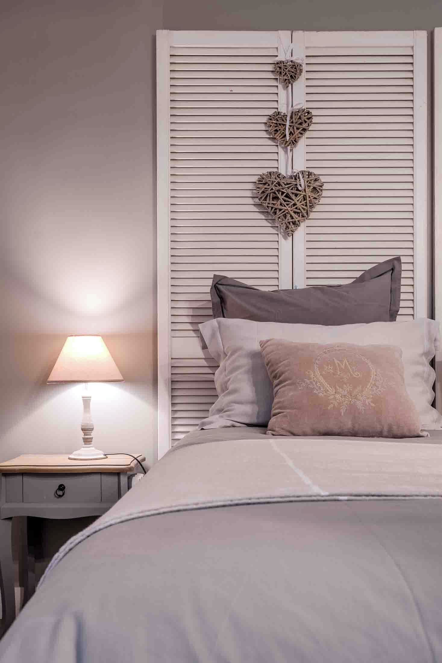 Cama bem arrumada com edredom branco, luminária de cabeceira e mesa de apoio com um vaso.