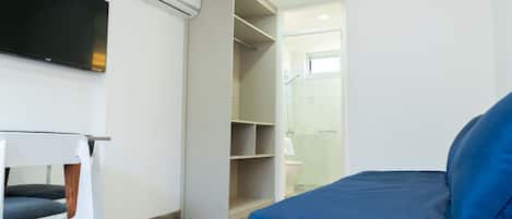 Quarto de hotel com cama, televisão fixada na parede, uma cadeira e banheiro visível por uma porta aberta.