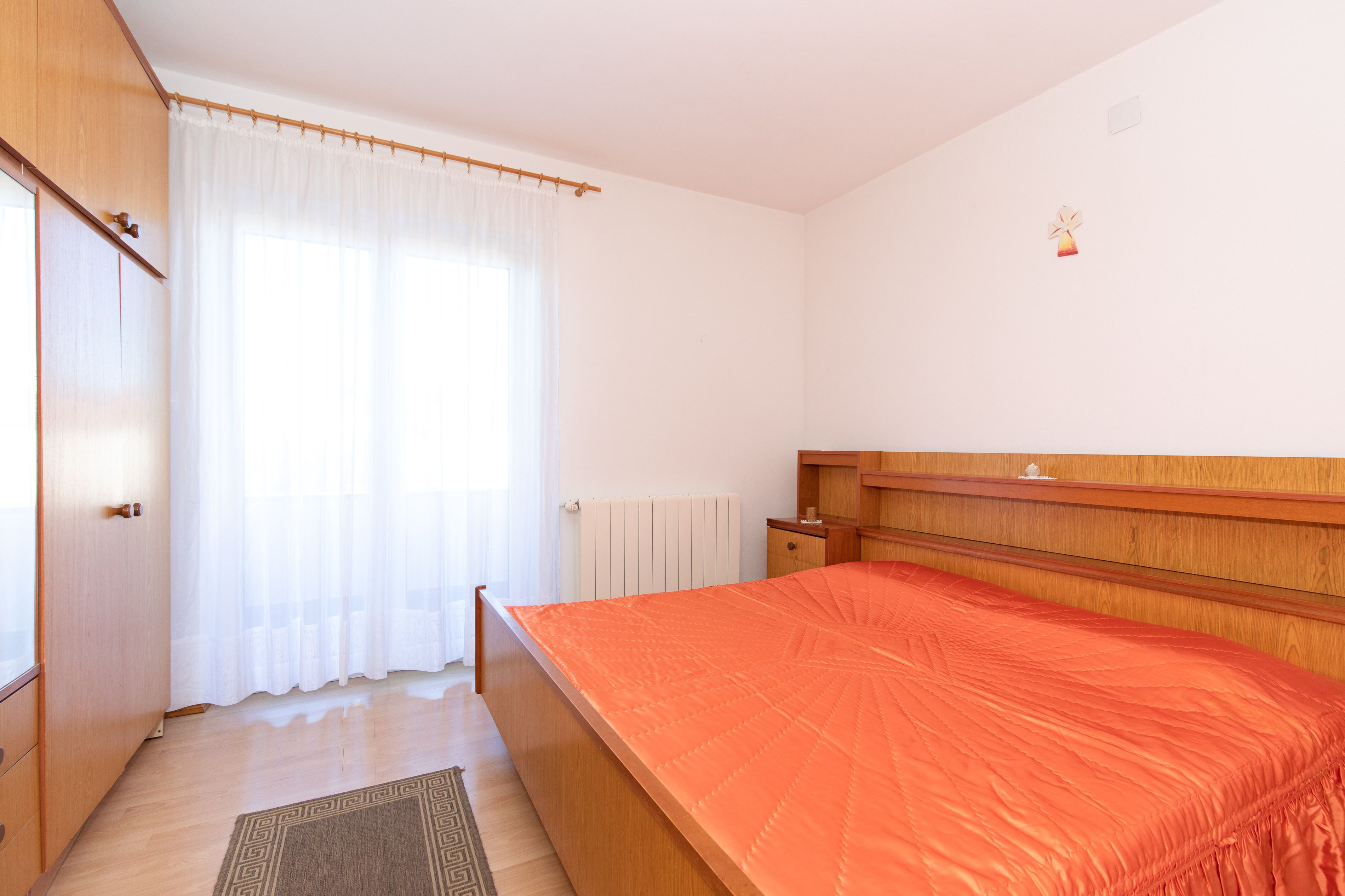 Quarto com cama de madeira, radiador branco e janela com cortinas finas.