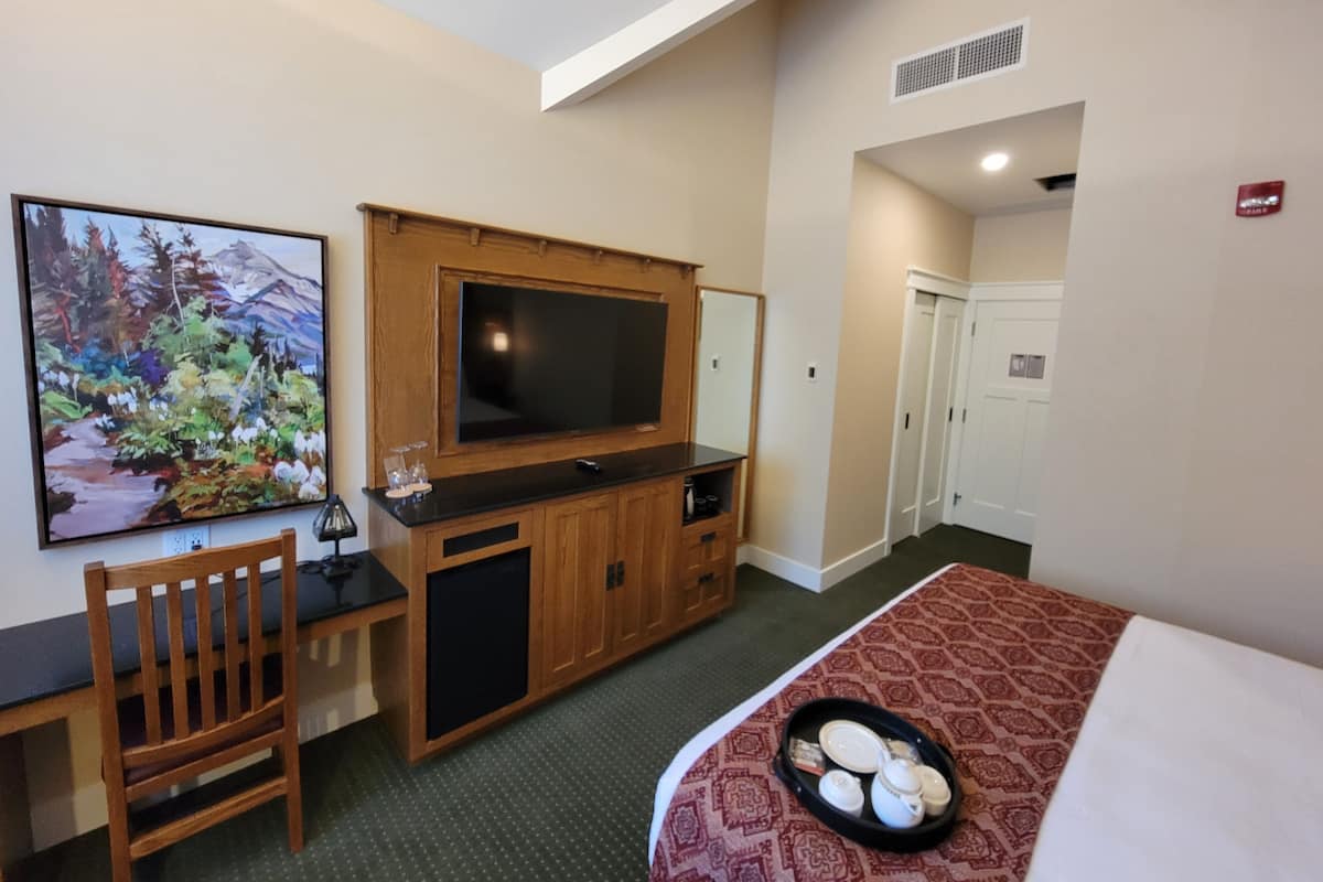 Quarto de hotel com uma cama, uma escrivaninha com cadeira, uma televisão presa a um armário de madeira e um quadro na parede.