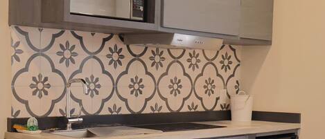 Cozinha compacta com armários cinza, micro-ondas e uma pequena geladeira. Apresenta um painel decorativo e uma mesa redonda de madeira com banquinhos.