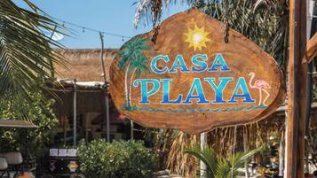 Uma placa de madeira com a inscrição 'CASA PLAYA', emoldurada por palmeiras e uma construção com teto de palha.
