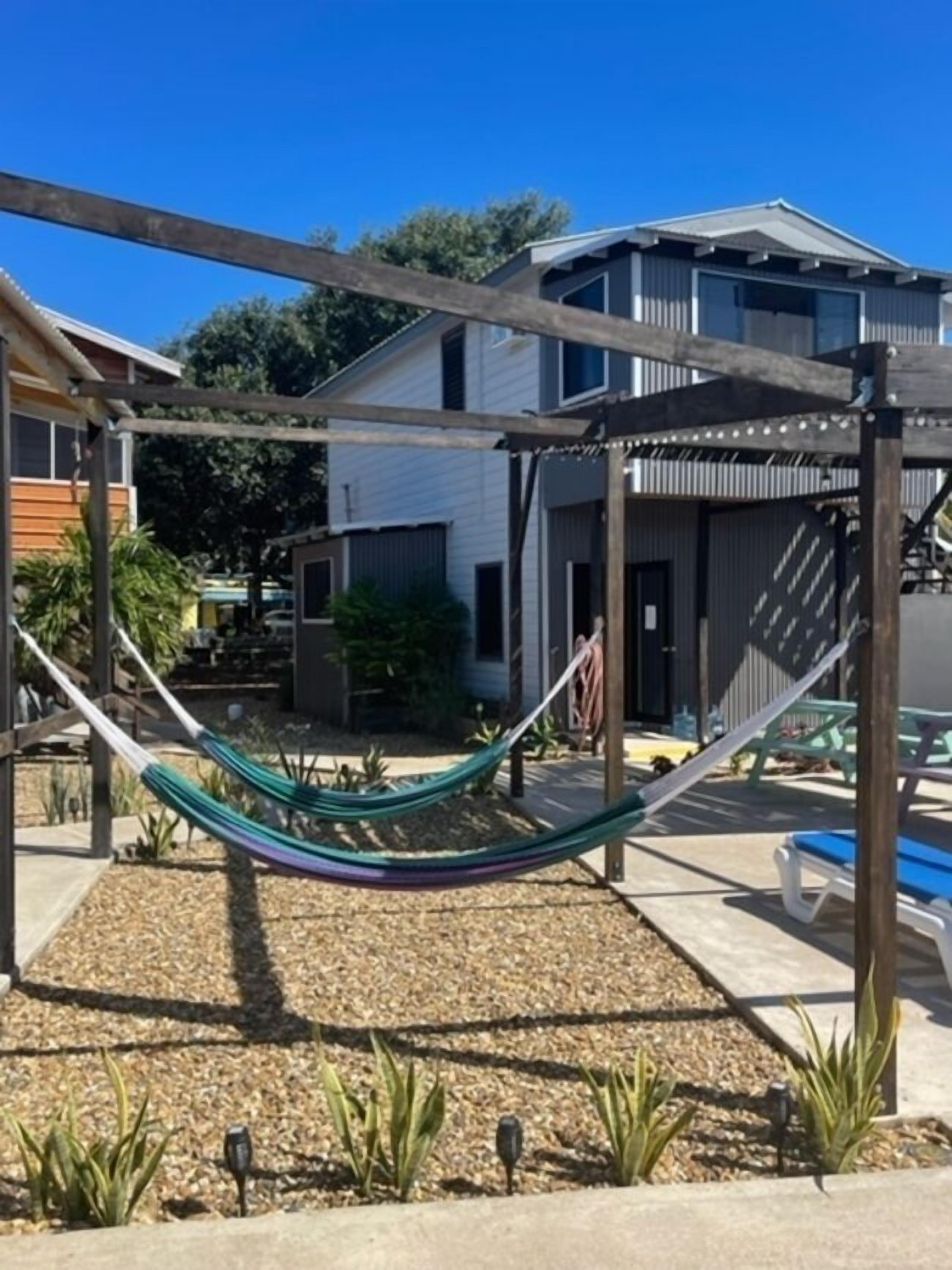 Uma rede sob uma pérgola de madeira em um quintal com piscina e uma casa ao fundo.