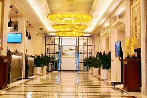 Um hall de hotel com piso de mármore, balcão de recepção, plantas em vasos e um grande candelabro.