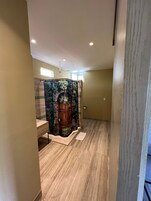 Uma casa de banho com toucador de madeira, chuveiro com parede revestida a azulejos verdes e castanhos, e porta de madeira.