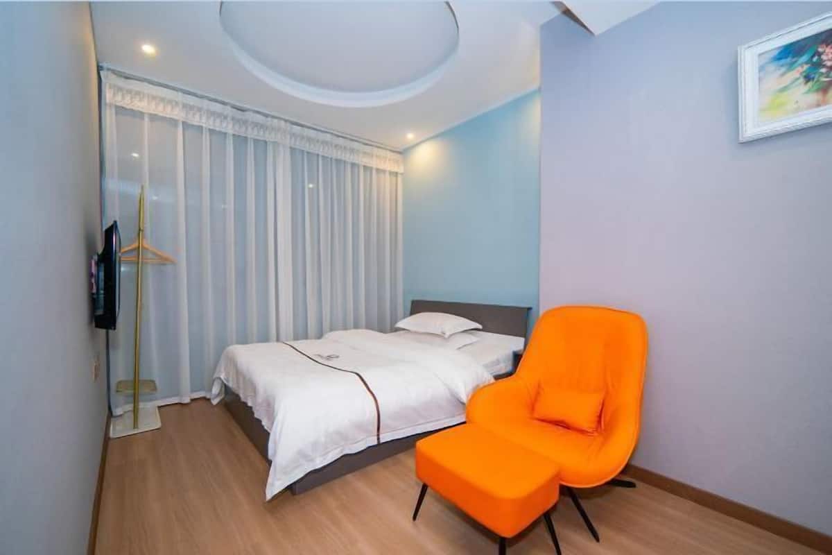 Quarto de hotel com cama, poltrona laranja, cabideiro e um quadro na parede.
