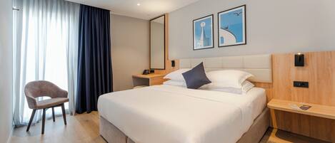 Quarto de hotel moderno com uma cama grande, mesa de cabeceira, uma cadeira e uma janela ampla com cortinas.