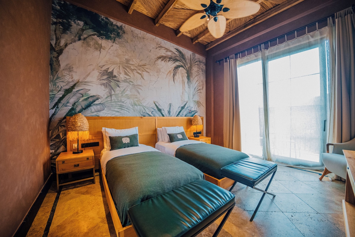 Quarto de hotel com duas camas, um mural grande na parede, um ventilador de teto e vista para o exterior pela janela.