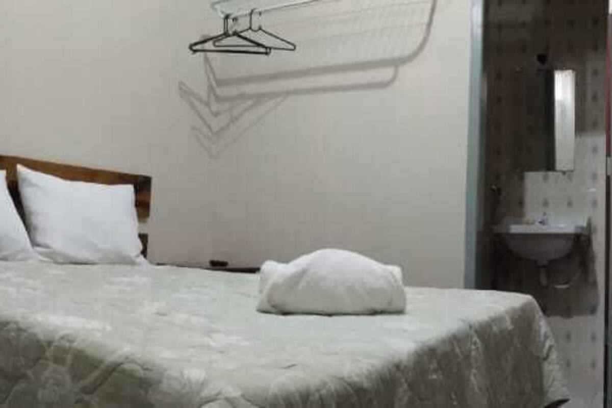 Quarto com cama, travesseiros, cabeceira de madeira, mesa de cabeceira e cabides fixados na parede.