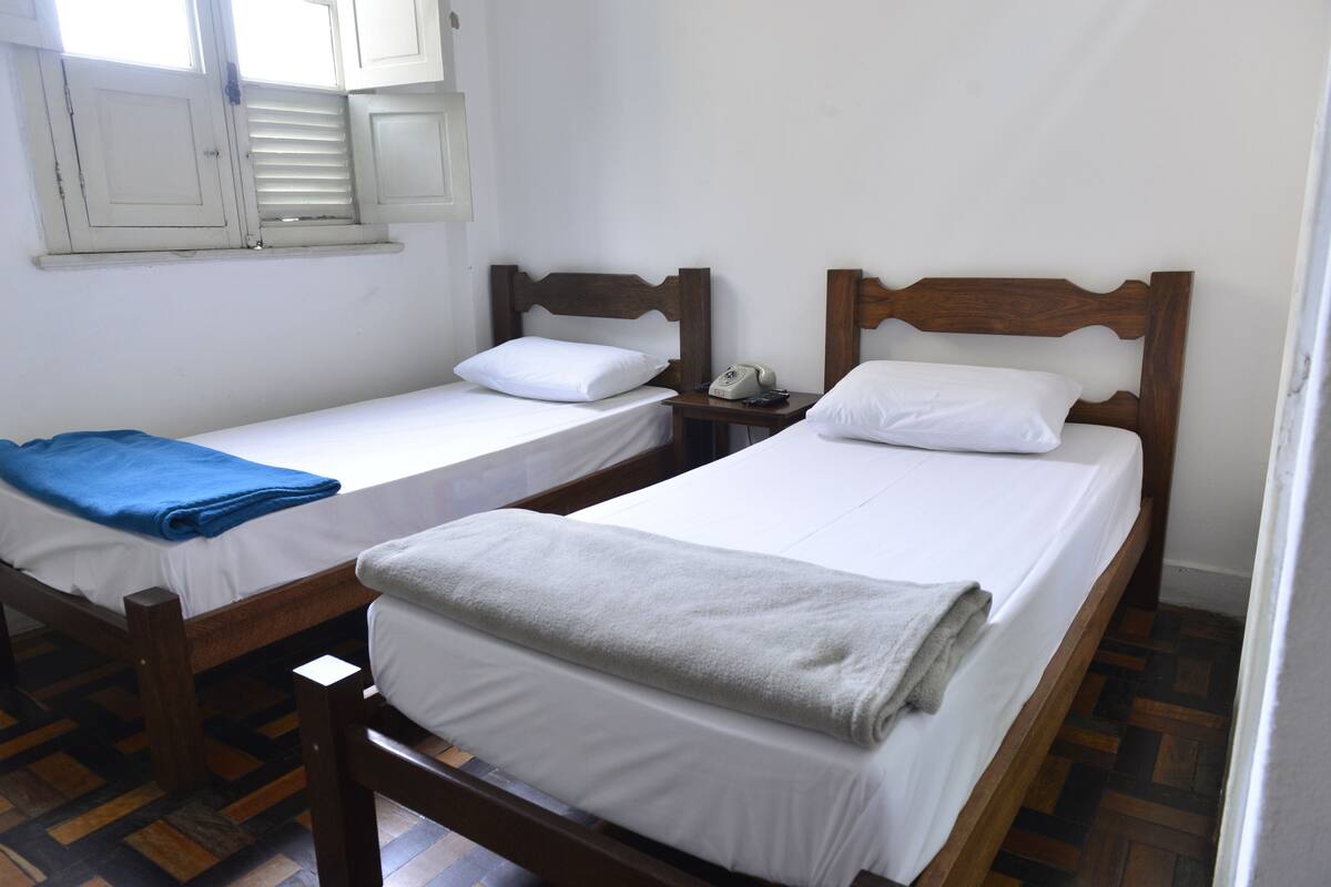 Duas camas de solteiro com estruturas de madeira, roupa de cama branca e uma pequena mesa de cabeceira com telefone.