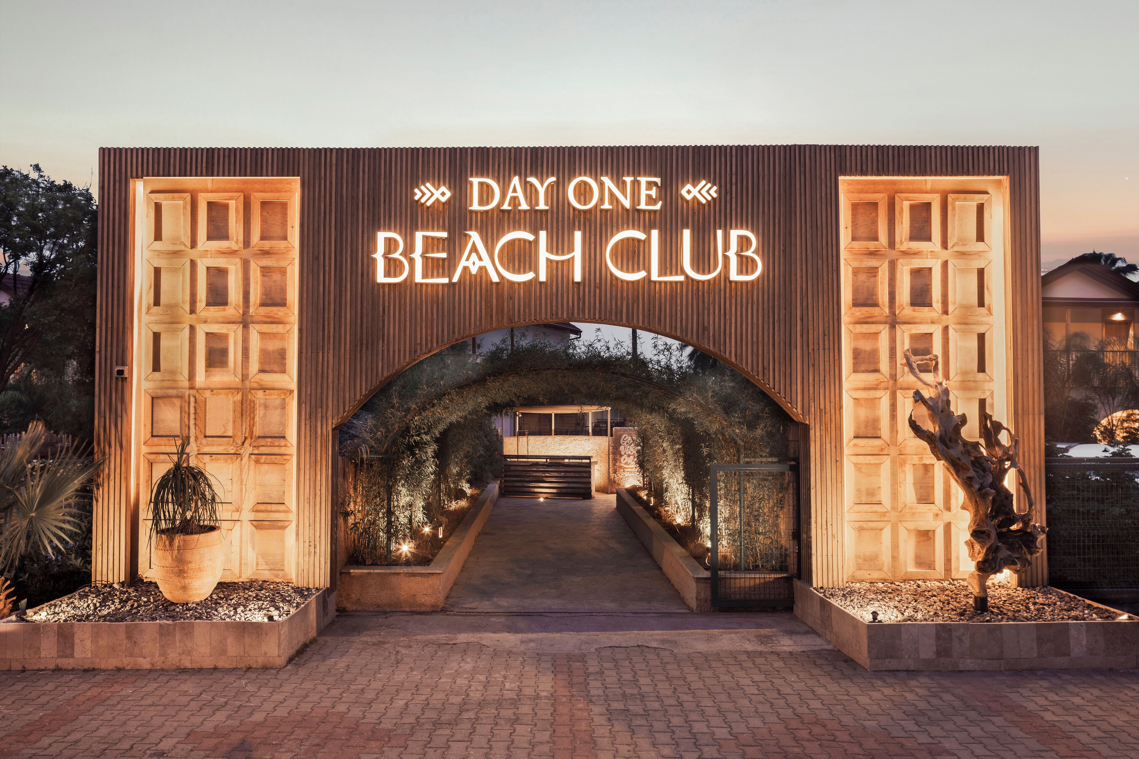 A entrada do Day One Beach Club, com fachada de madeira e sinalização iluminada.