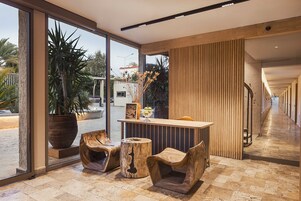 Uma área de recepção moderna com um balcão de madeira, duas cadeiras de madeira, uma grande planta em vaso e vista para o exterior.