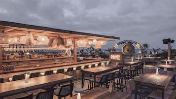 Uma área de bar ao ar livre com mesas e cadeiras de madeira, balcão de bar com garrafas e luminárias pendentes.