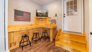 Uma área de bar com balcão de madeira, banquetas, uma pintura de paisagem e uma bandeira americana.