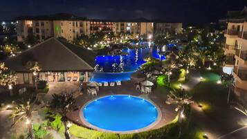 Resort com uma piscina central ampla, piscinas menores ao redor, e área externa bem iluminada com espreguiçadeiras e guarda-sóis.