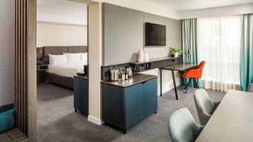 Quarto de hotel com cama, escrivaninha com cadeira, televisão e janela com cortinas.