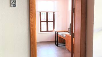 Quarto com porta de madeira, janela com grades, cama e uma pequena mesa.