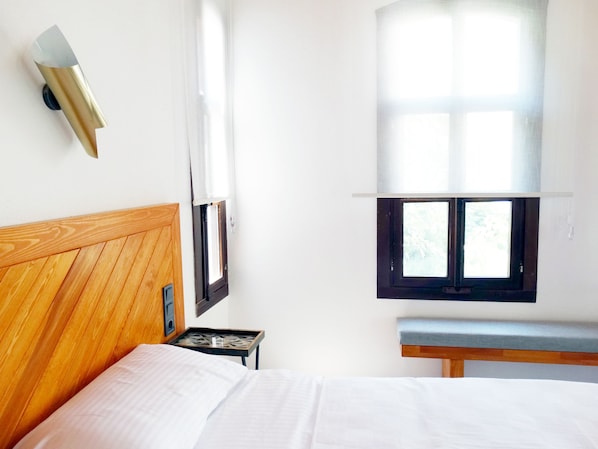 Quarto com cabeceira de madeira, mesa de cabeceira com abajur, janela com vista e luminária na parede.