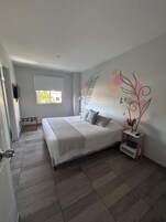 Quarto com cama, mesa de cabeceira, televisão na parede, janela e porta.