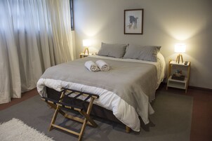 Cama bem arrumada com toalhas enroladas, duas mesas de cabeceira com abajures, um quadro na parede e um banco de madeira em frente à cama.