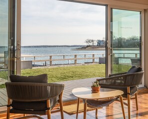 Uma área de estar moderna ao ar livre com duas cadeiras de vime e uma mesa pequena, com vista para a água e um deck de madeira.