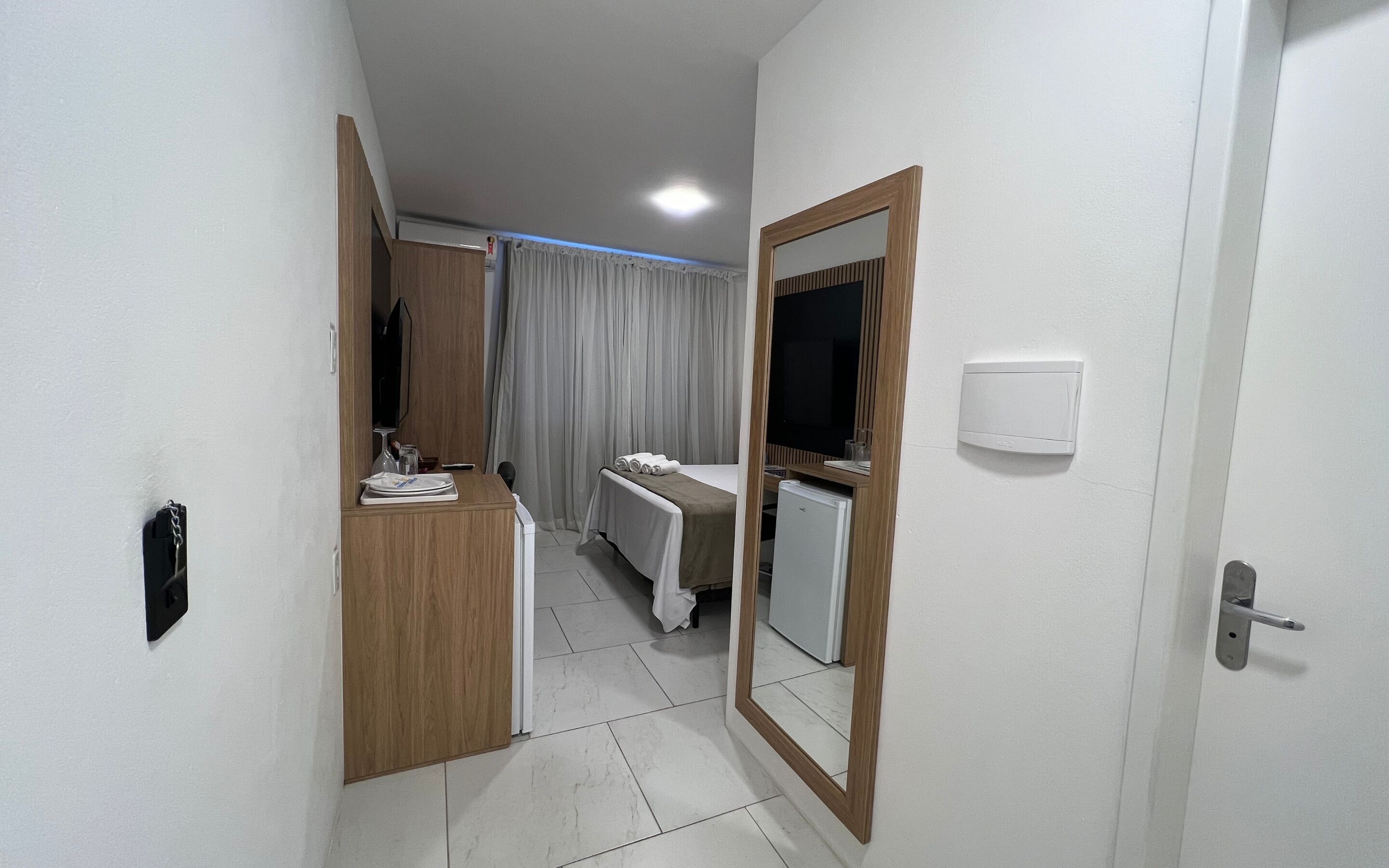 Quarto de hotel com cama, escrivaninha, cadeira, televisão e um pequeno frigorífico.