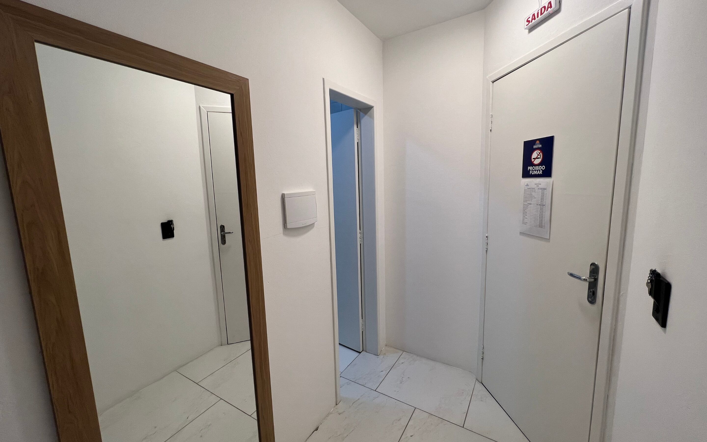 Um corredor com paredes brancas, uma porta de moldura em madeira e uma porta de moldura azul. Há uma placa acima da porta azul com a palavra 'SAIDA' e outra placa que proíbe fumar.