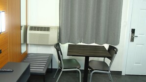 Quarto pequeno com uma mesa, duas cadeiras, janela com cortina, ar condicionado e uma porta.