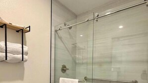 Uma casa de banho com chuveiro em cabine de vidro, sanita branca e porta-toalhas com toalhas dobradas.