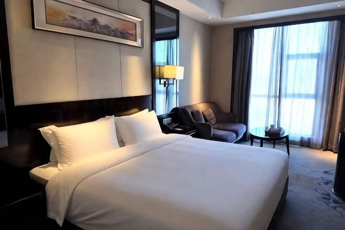 Quarto de hotel com uma cama grande, uma cadeira, uma mesinha e uma janela com cortinas.