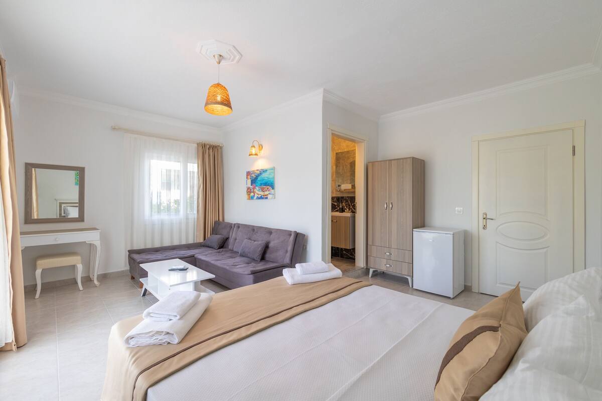 Quarto de hotel com cama, sofá, escrivaninha, guarda-roupa e uma pequena cozinha.