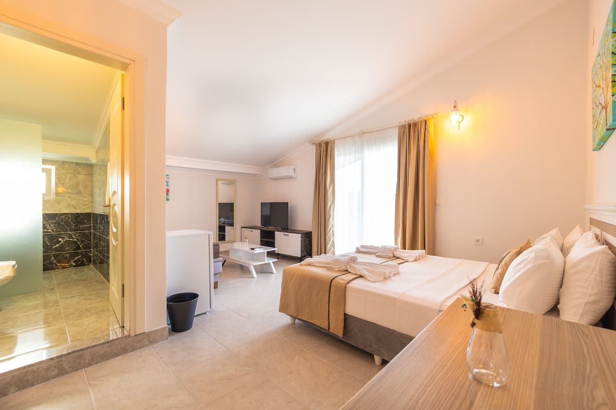 Quarto de hotel com cama, mesa de cabeceira e escrivaninha com cadeira. Inclui banheiro com vaso sanitário, pia e chuveiro. O quarto possui uma janela grande com cortinas.